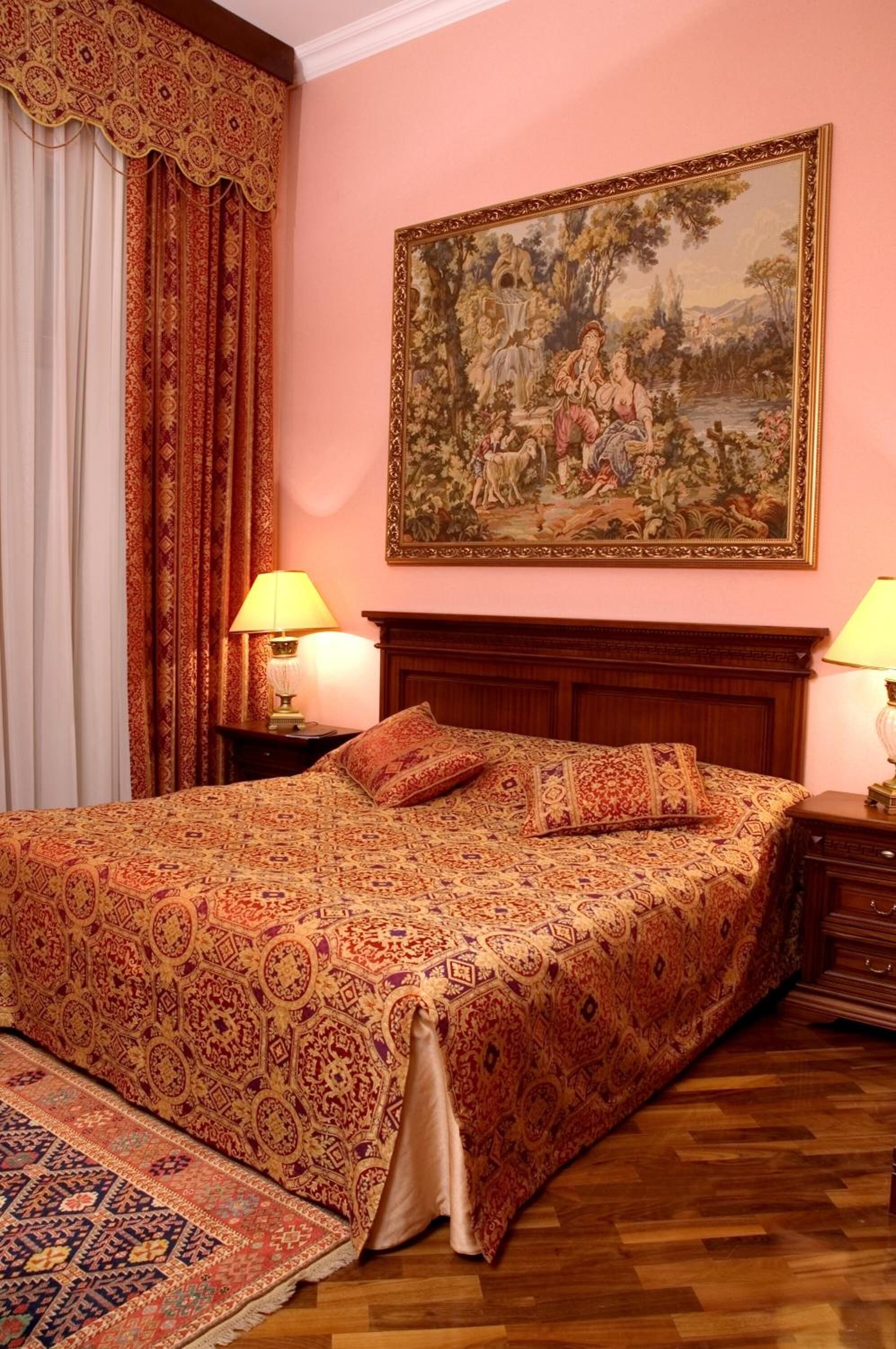 Uma cama grande com colcha estampada, duas luminárias de cabeceira, um quadro sobre a cama e piso de madeira.