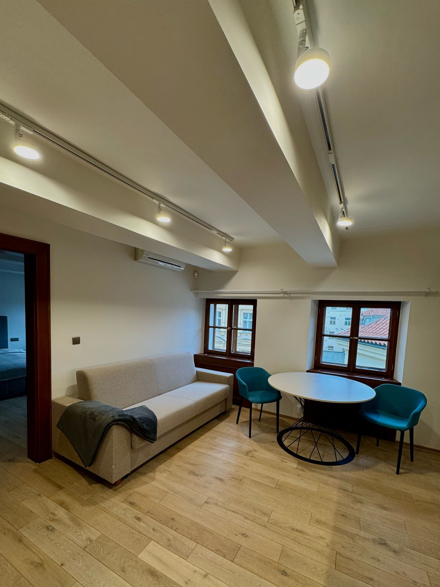 Quarto com sofá, mesa redonda e duas cadeiras. Janelas oferecem vista para edifícios.