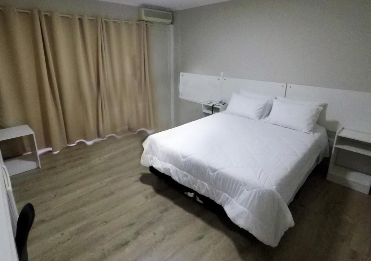 Quarto de hotel com uma cama grande, uma mesa de cabeceira, uma cadeira e uma cortina.