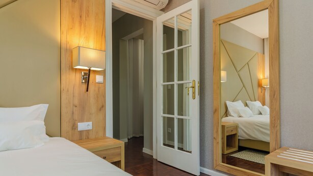 Quarto de hotel com parede de painel de madeira, cama com lençóis brancos, mesa de cabeceira, espelho e porta que dá para outro quarto.