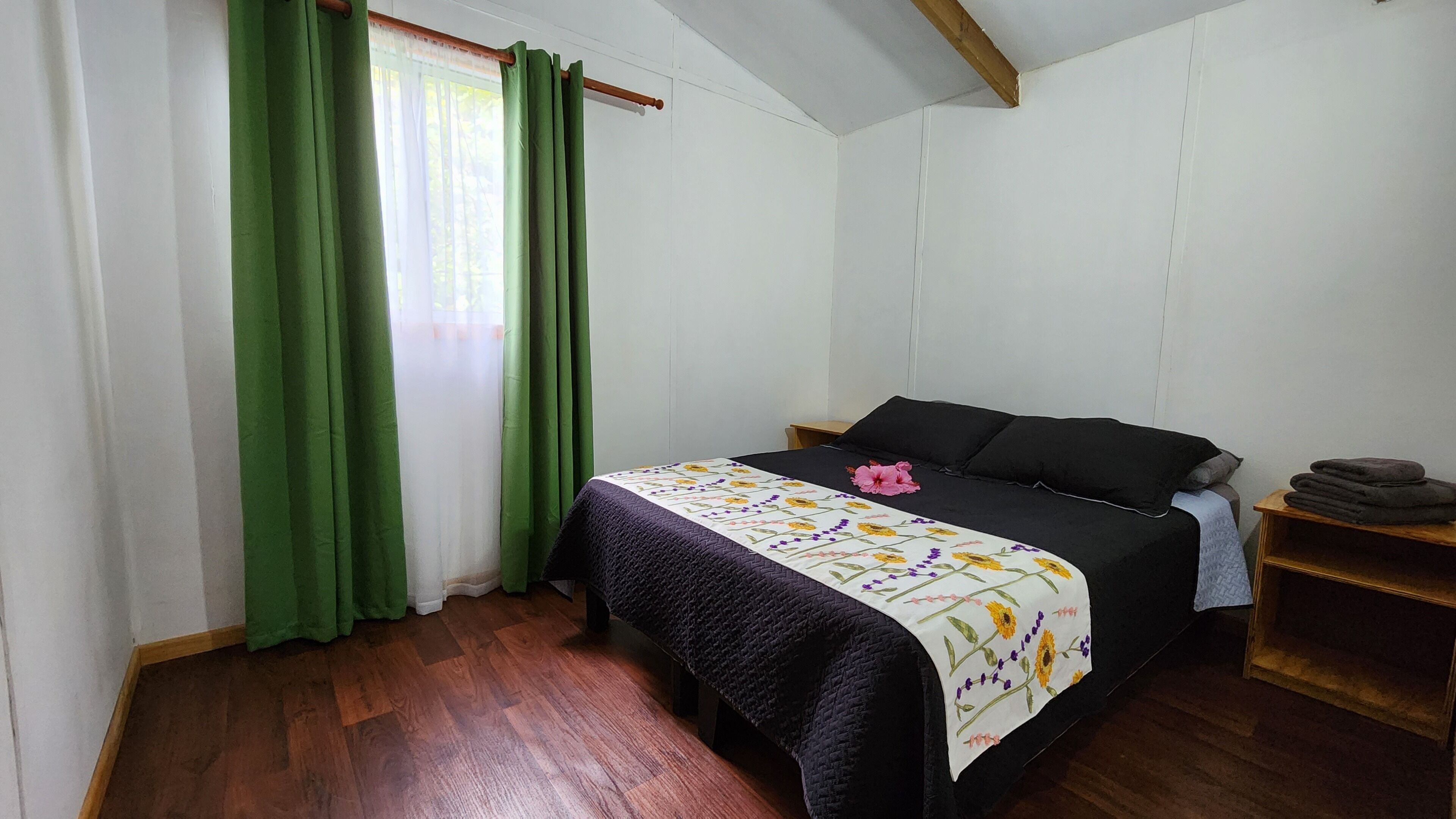 Quarto com cama, cortinas verdes, piso de madeira e mesa de cabeceira com um cobertor dobrado.