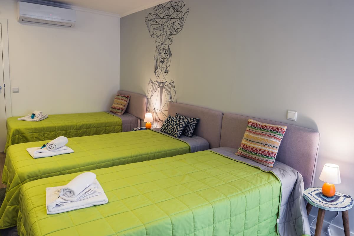 Quarto de hotel com duas camas de solteiro, uma obra de arte na parede, uma mesinha com abajur e uma janela.