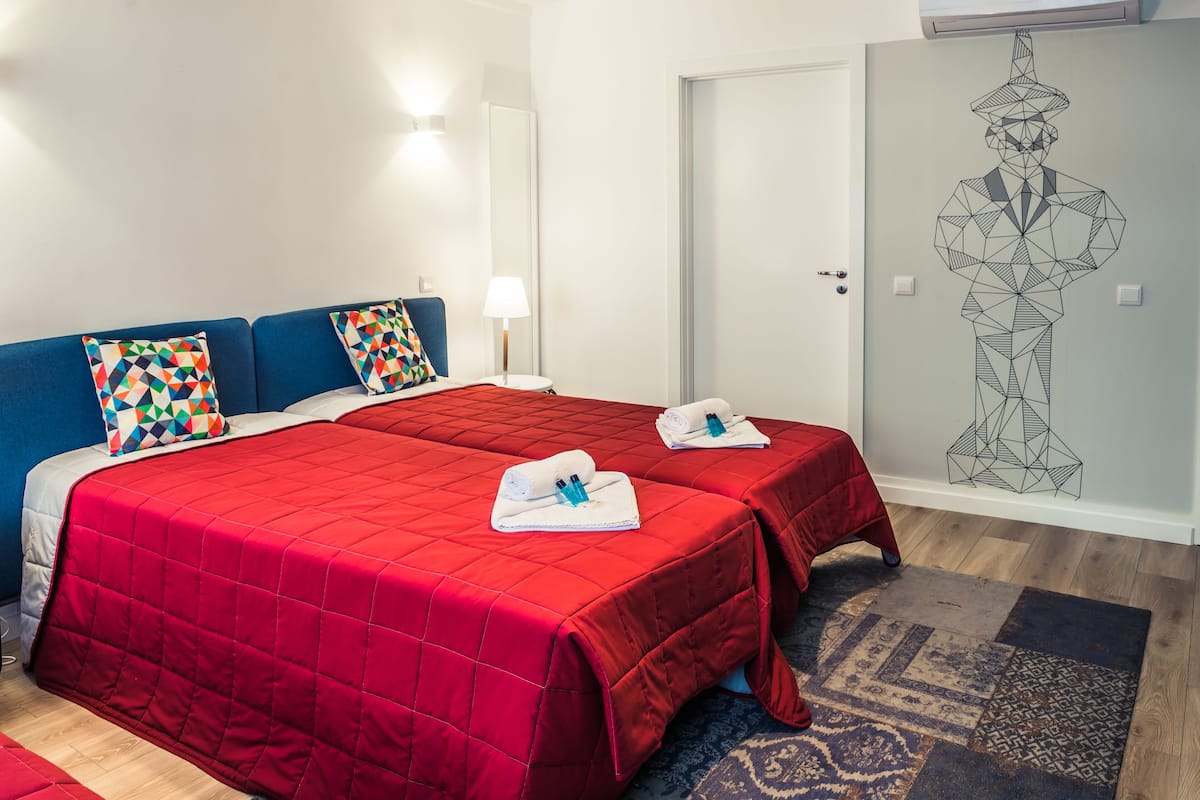 Quarto de hotel com duas camas, cada uma com colcha acolchoada vermelha e almofadas geométricas coloridas. Há uma unidade de ar condicionado na parede acima das camas, uma porta com adesivo geométrico na parede e um tapete estampado sobre o chão de madeira.