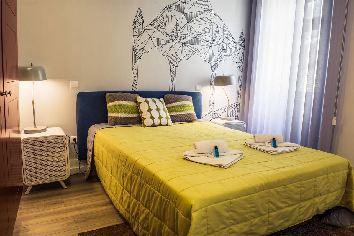 Quarto de hotel com cama, arte geométrica na parede, ventoinha e janela com cortinas.
