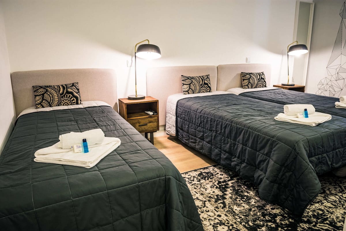 Quarto de hotel com três camas, cada uma com um edredom verde escuro, uma luminária de cabeceira e uma toalha dobrada com uma garrafa em cima.