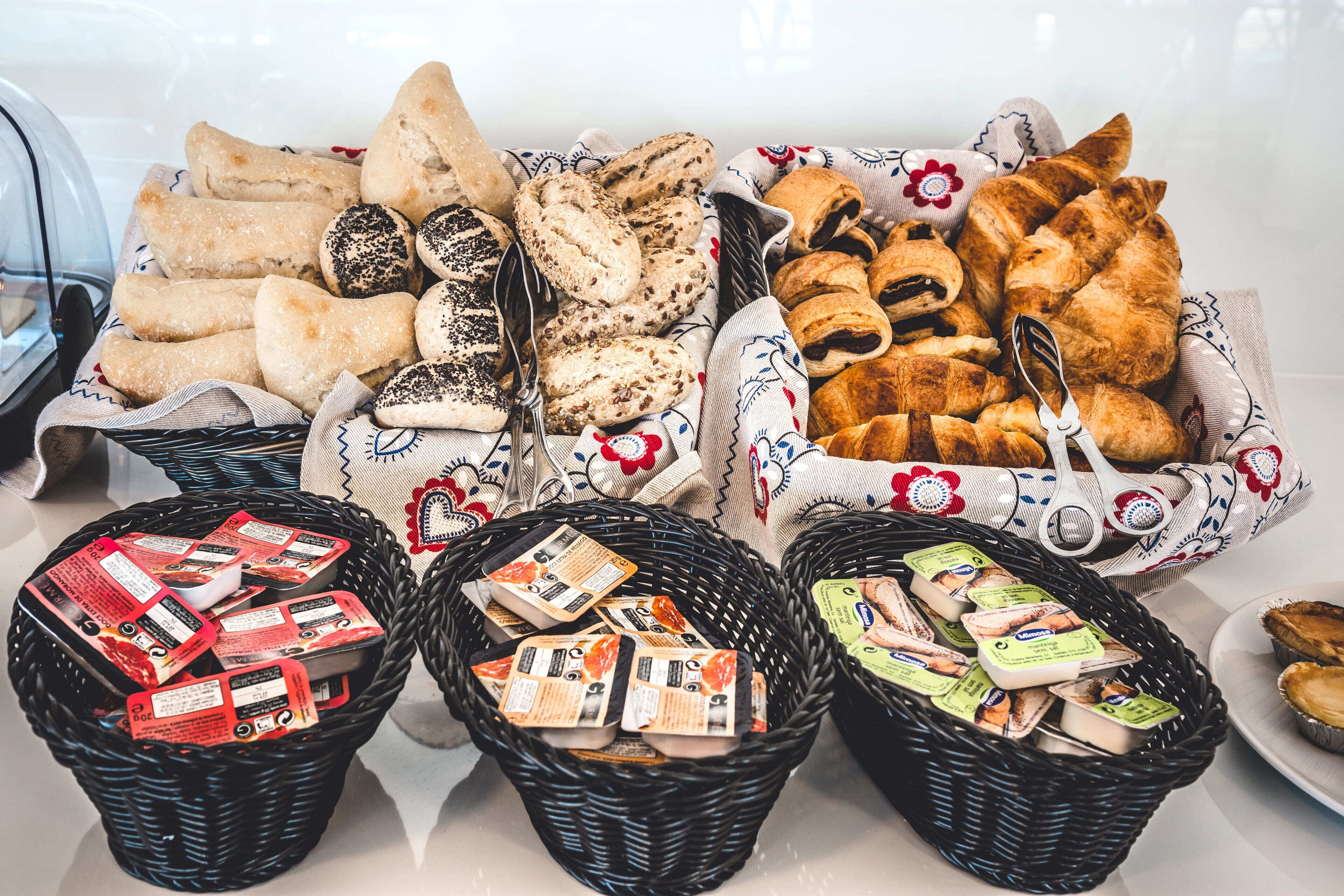 Diversos produtos de panificação expostos em cestas e sobre uma mesa, incluindo pão, pães de forma e doces.