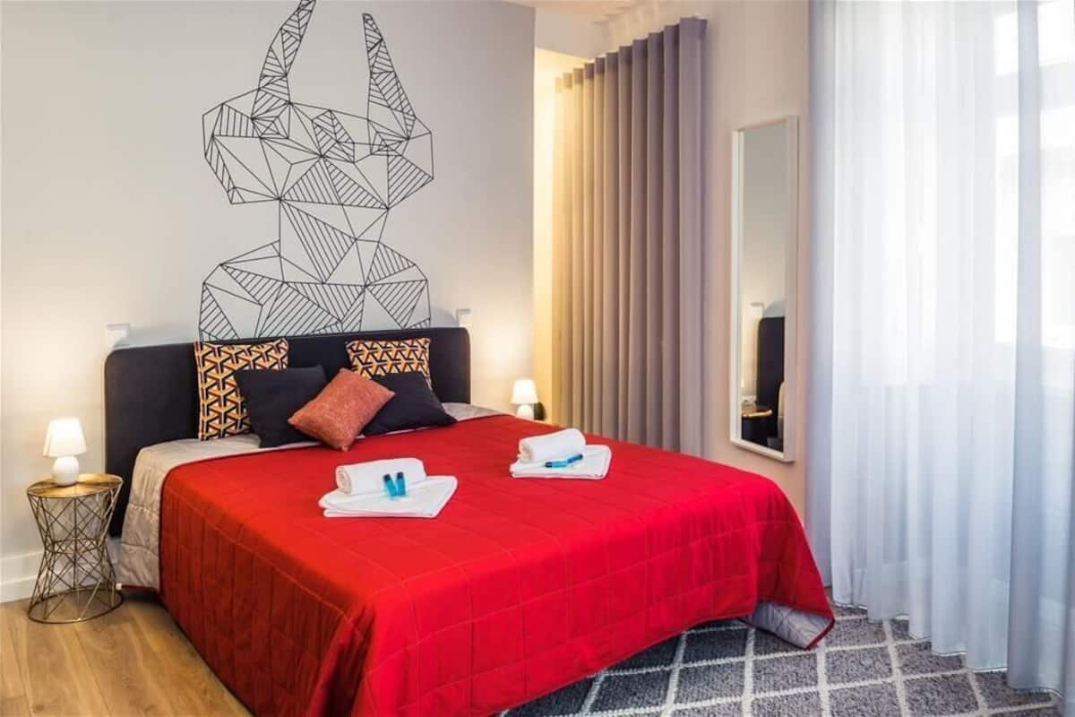 Quarto de hotel com colcha vermelha, arte geométrica na parede, uma janela grande com cortinas e um abajur.