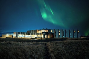 Um edifício moderno com telhado plano e amplas janelas, situado numa área gramada com vista para o céu noturno e a aurora boreal.
