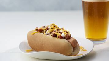 Um cachorro-quente com chili e mostarda no pão, colocado em um prato branco, com um copo de cerveja ao fundo.