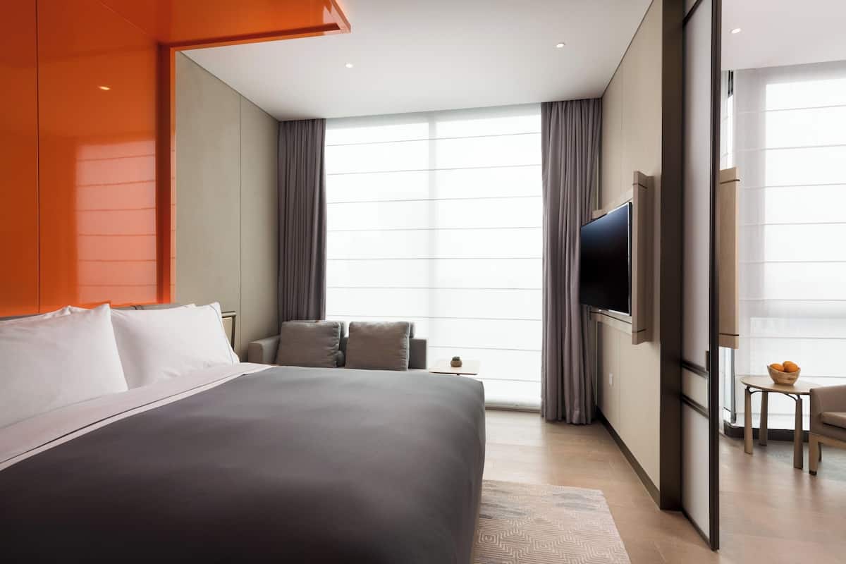 Quarto de hotel moderno com uma cama grande, televisão, uma mesinha com frutas e vista para a cidade.