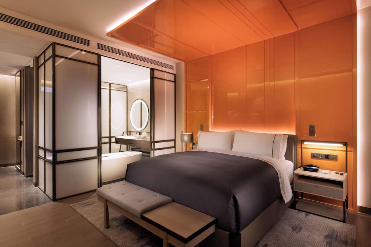 Quarto de hotel moderno com uma cama grande, mesa de cabeceira e um banco.