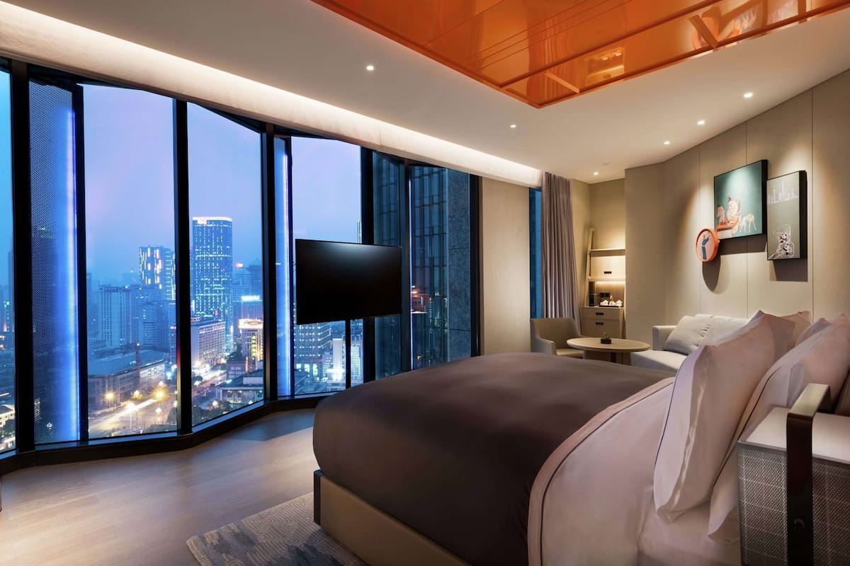 Quarto de hotel moderno com uma cama grande, televisão, escrivaninha e vista para a cidade.
