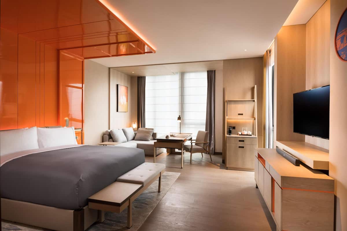 Quarto de hotel moderno com cama grande, sofá, área de refeições, TV e pequena cozinha.