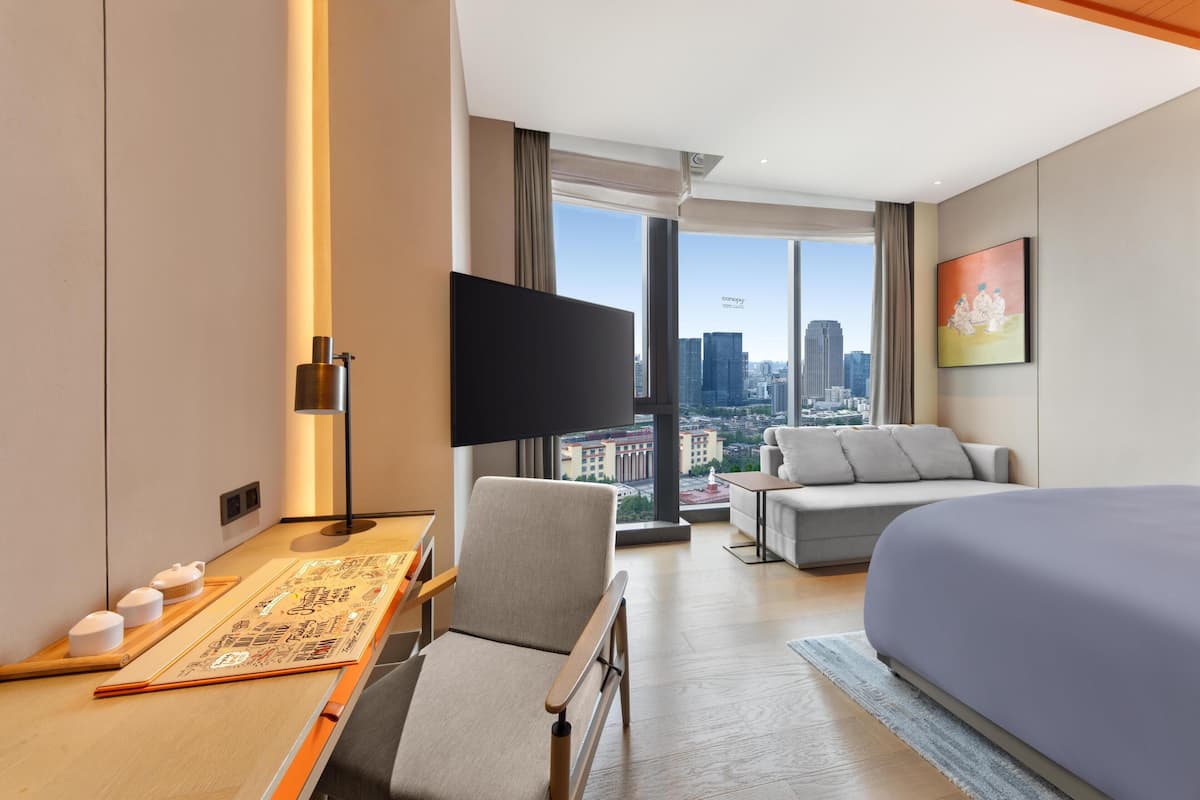 Quarto de hotel com cama, escrivaninha com televisão de tela plana, sofá e grande janela com vista para a cidade.