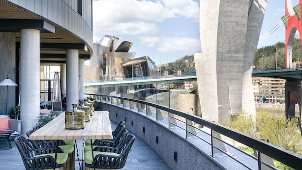 Área externa com mesas e cadeiras, com vista para um rio e uma ponte.