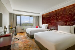 Quarto de hotel com duas camas, uma escrivaninha, uma cadeira, uma mesinha e uma janela grande com vista para a cidade.