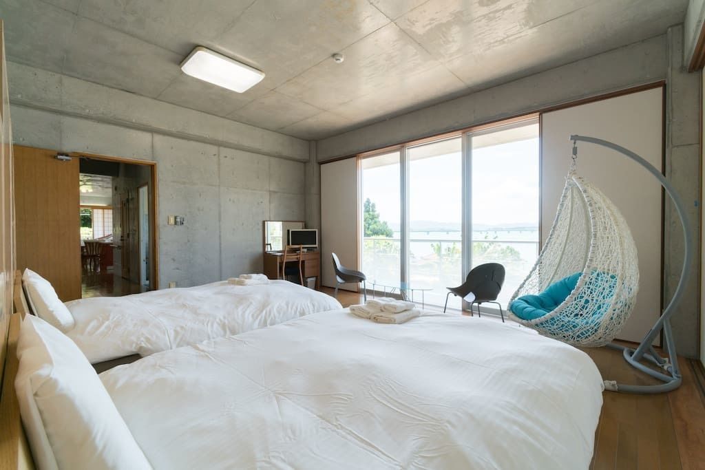 Quarto de hotel com duas camas, uma escrivaninha, uma cadeira e uma janela grande com vista.