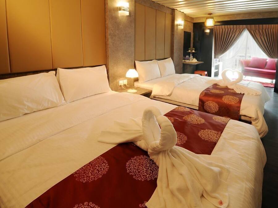 Quarto de hotel com duas camas, cada uma com roupa de cama branca e colcha vermelha estampada. Há luminárias laterais e uma janela com cortinas.