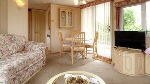 Uma sala de estar com sofá estampado, mesa de jantar com cadeiras, televisão em um móvel e ventilador de teto.