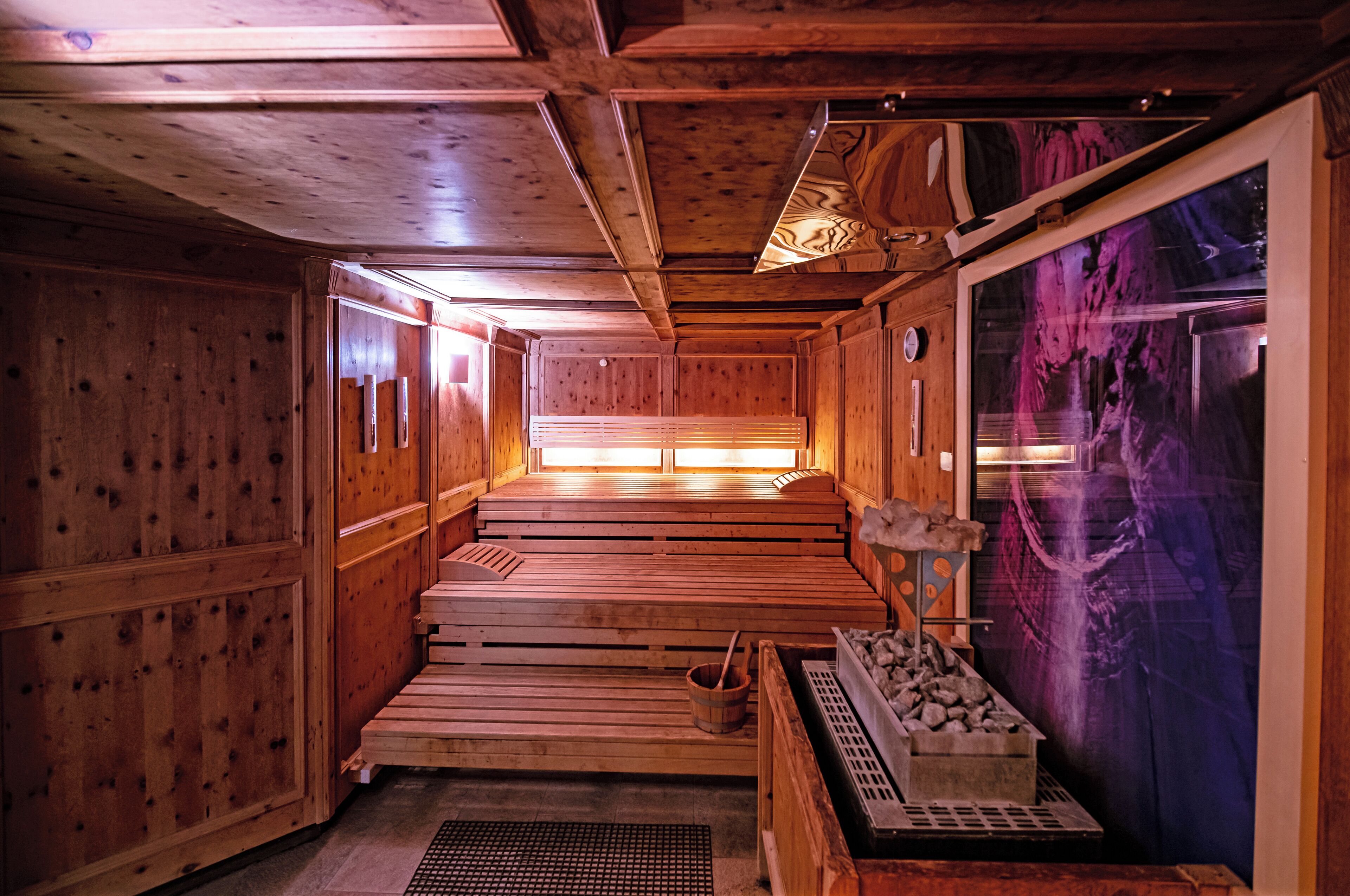 Uma sauna de madeira com bancos, uma bacia de pedra e uma grande janela com vista para uma montanha.