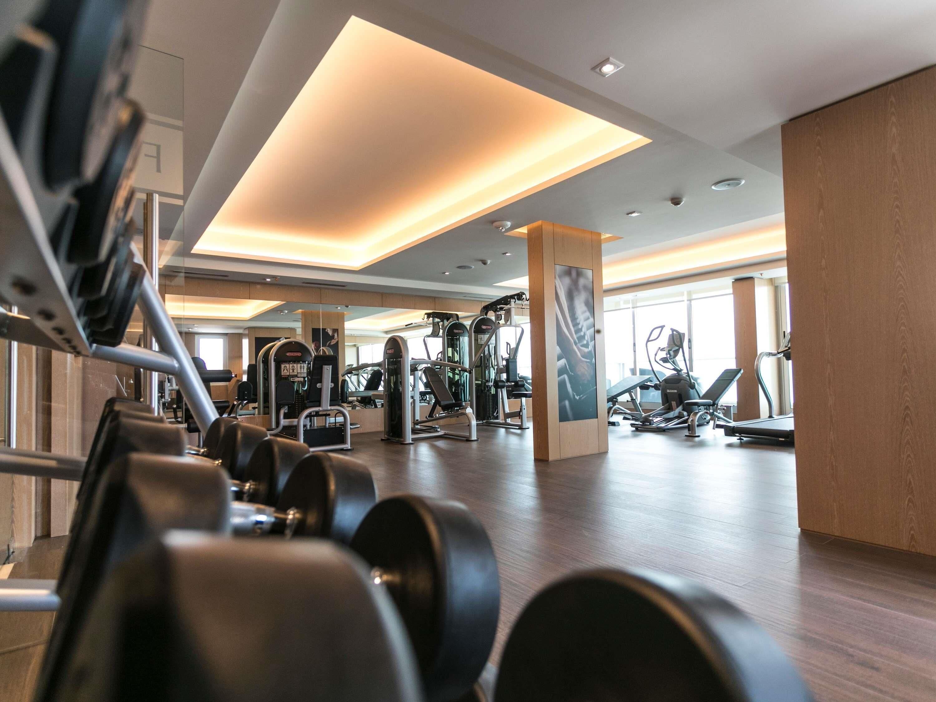 Uma academia com diversos equipamentos de ginástica, piso de madeira e amplas janelas que permitem a entrada de luz natural.