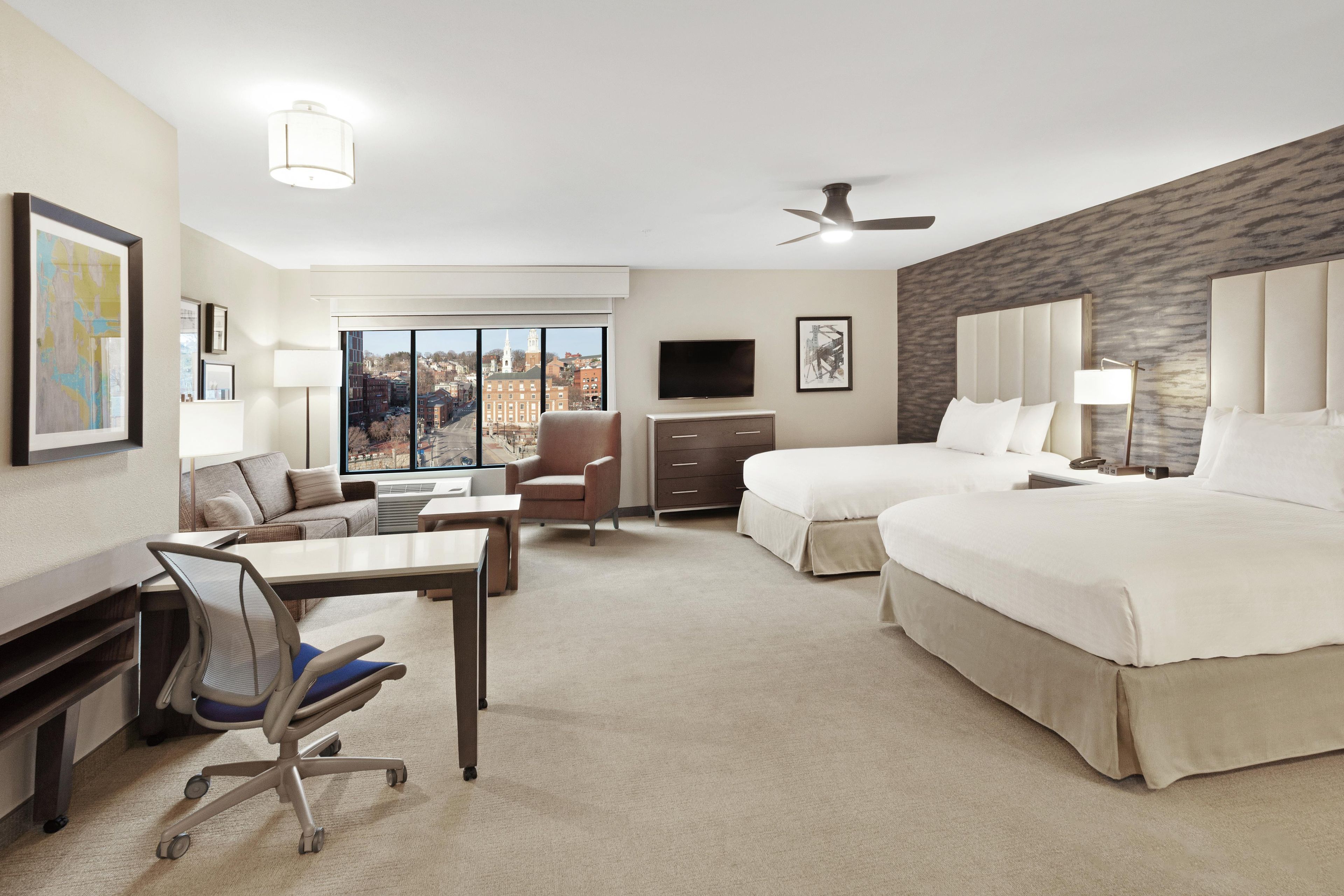 Quarto de hotel com uma escrivaninha, duas camas, uma cadeira, uma televisão e uma janela grande com vista para a cidade.