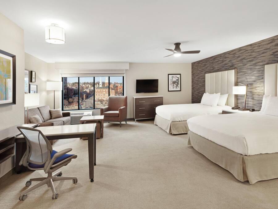 Quarto de hotel com uma escrivaninha, duas camas, uma cadeira, uma televisão e uma janela grande com vista para a cidade.