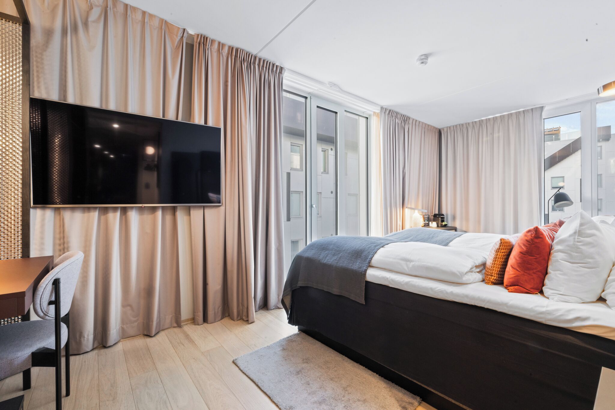 Quarto de hotel moderno com uma cama grande, televisão de tela plana na parede, escrivaninha e uma janela ampla com cortinas.