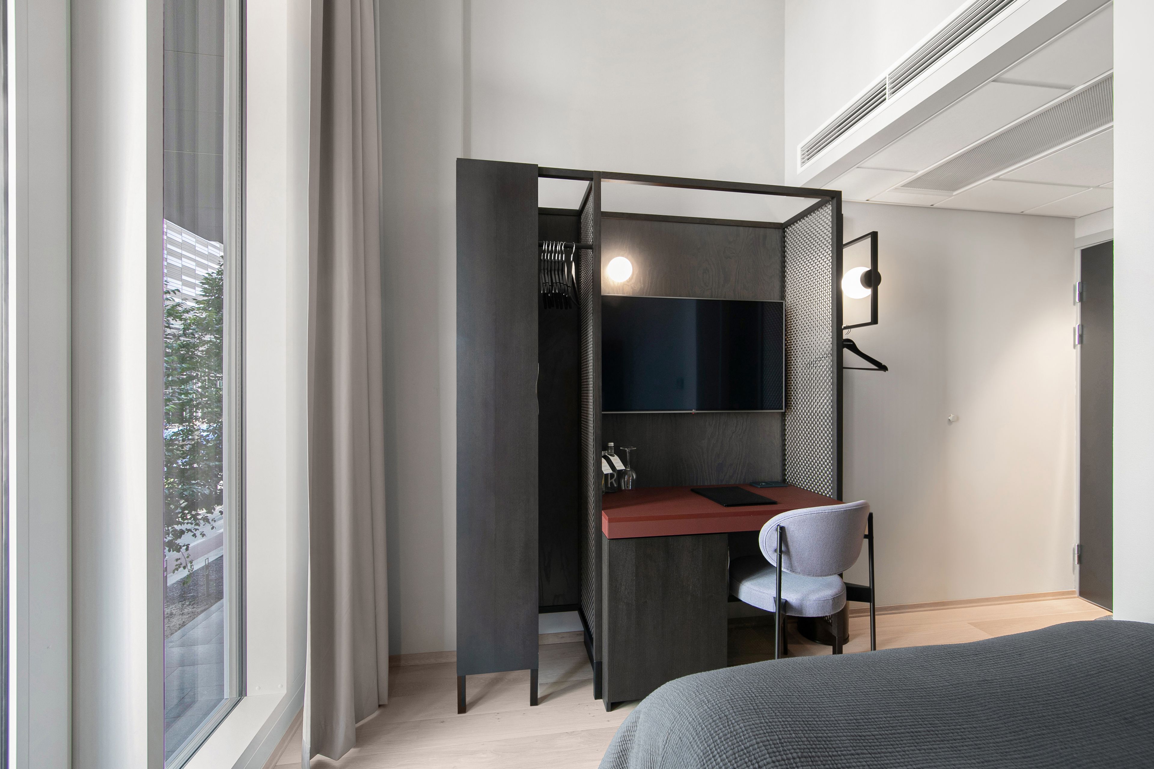 Quarto de hotel moderno com uma janela grande, uma mesa com cadeira, uma cama e um guarda-roupa.