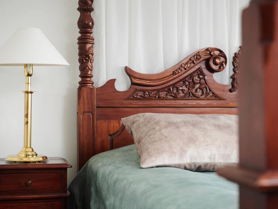 Uma cama de madeira com colcha verde e travesseiro branco, um abajur de latão e uma mesinha de cabeceira de madeira com uma gaveta.