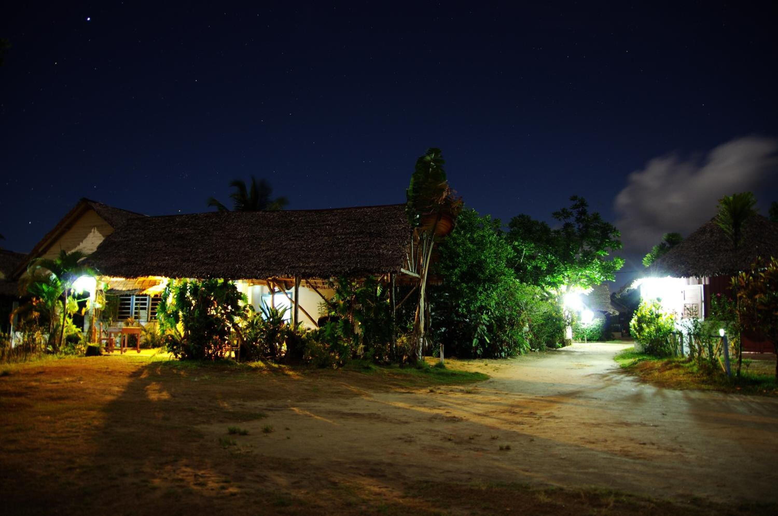 Paisagem rural com edifícios de telhado de palha, estrada de terra e vegetação ao redor.