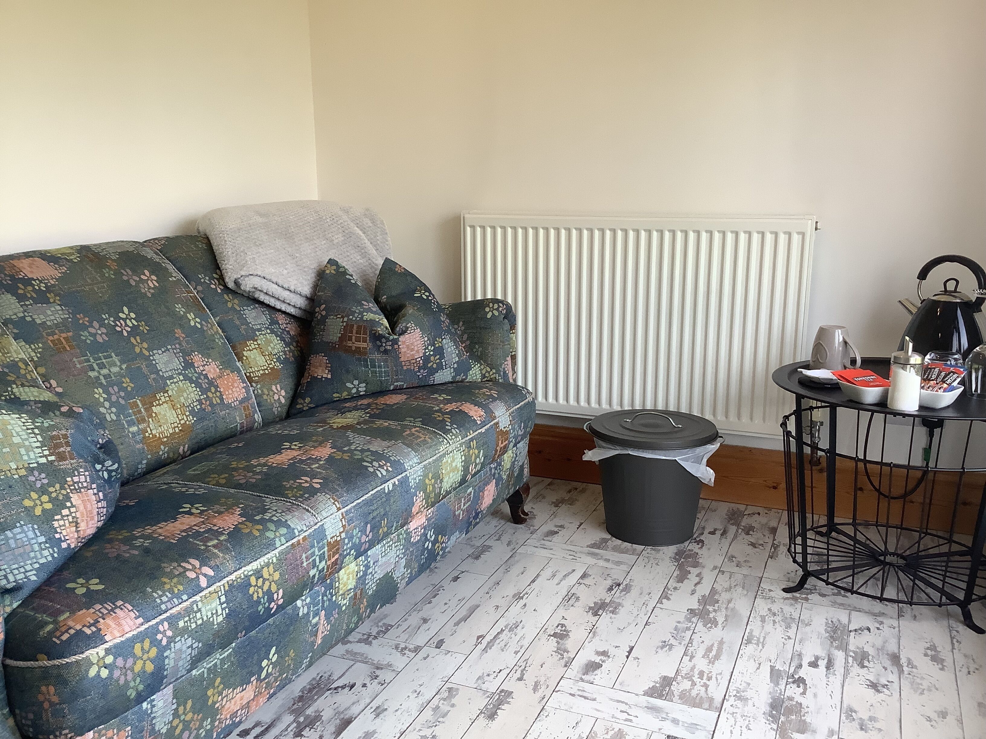 Uma sala de estar com um sofá estampado, um radiador, uma lixeira e uma mesa pequena com uma chaleira e xícaras.