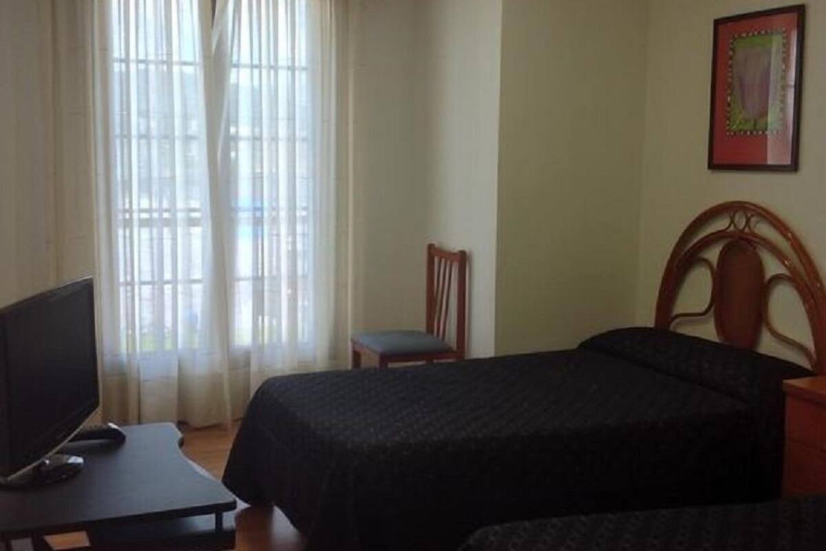 Quarto de hotel com duas camas, uma escrivaninha com computador, uma cadeira e uma janela com cortinas.