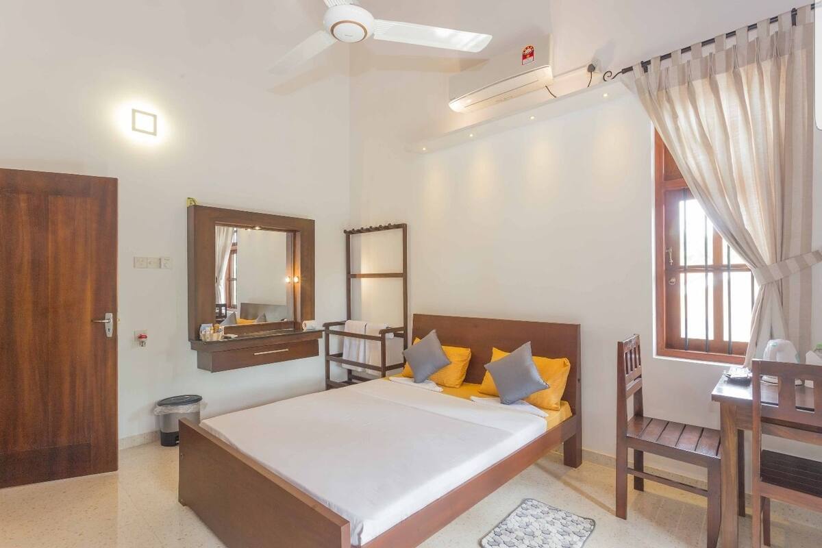 Quarto de hotel com uma cama, uma escrivaninha, uma cadeira, um espelho, um ventilador de teto e um ar condicionado.