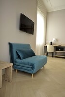 Quarto moderno com sofá verde-água, TV de tela plana na parede, pequena escrivaninha com luminária e janela com persianas.
