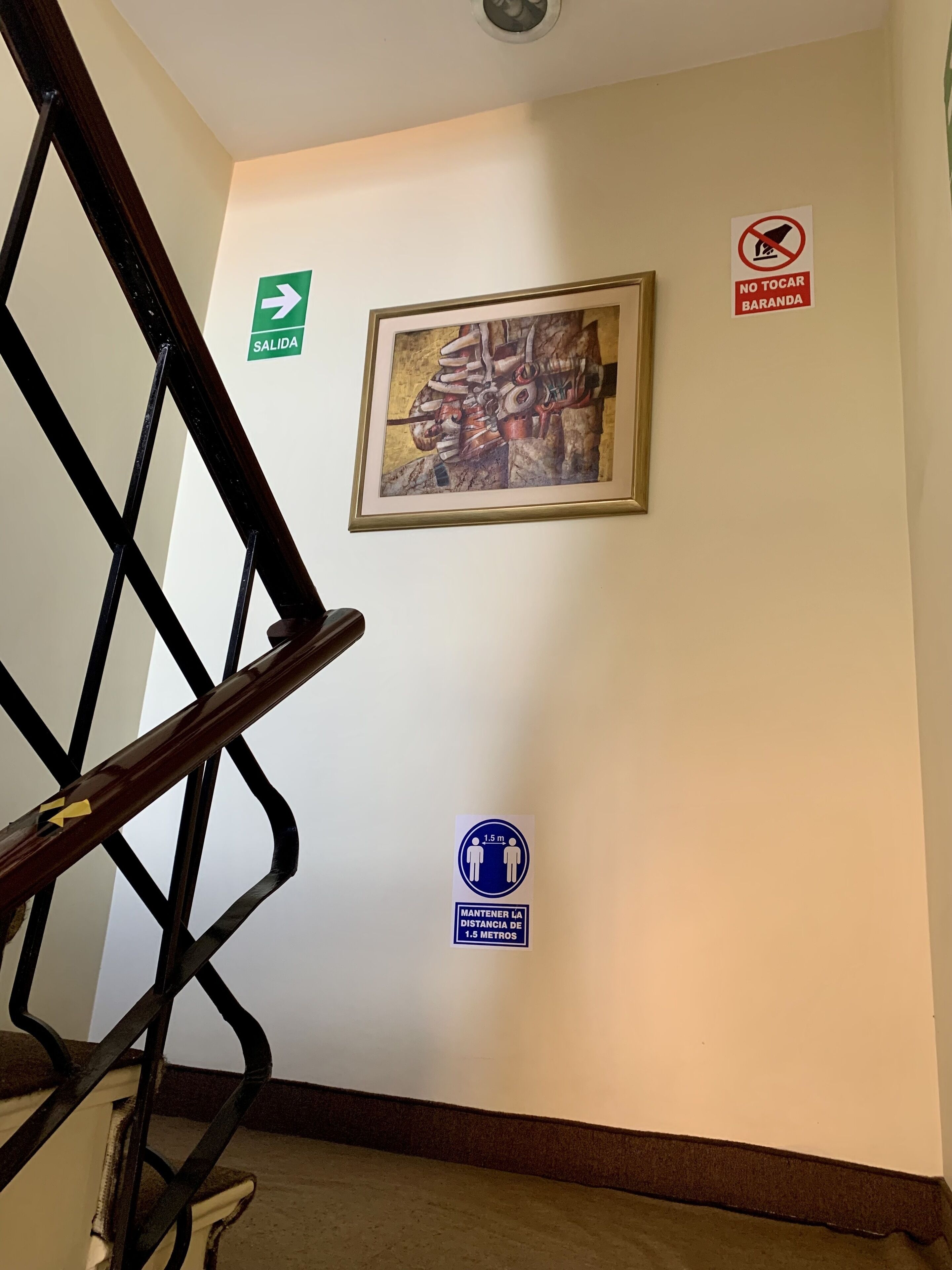 Um corredor com escadas, uma obra de arte emoldurada e placas de sinalização, incluindo uma saída e um aviso de distanciamento social.
