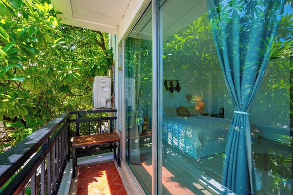 Varanda com mesa e cadeiras de madeira, porta de vidro que dá para um quarto com cama e cortinas, e vegetação exuberante do lado de fora.