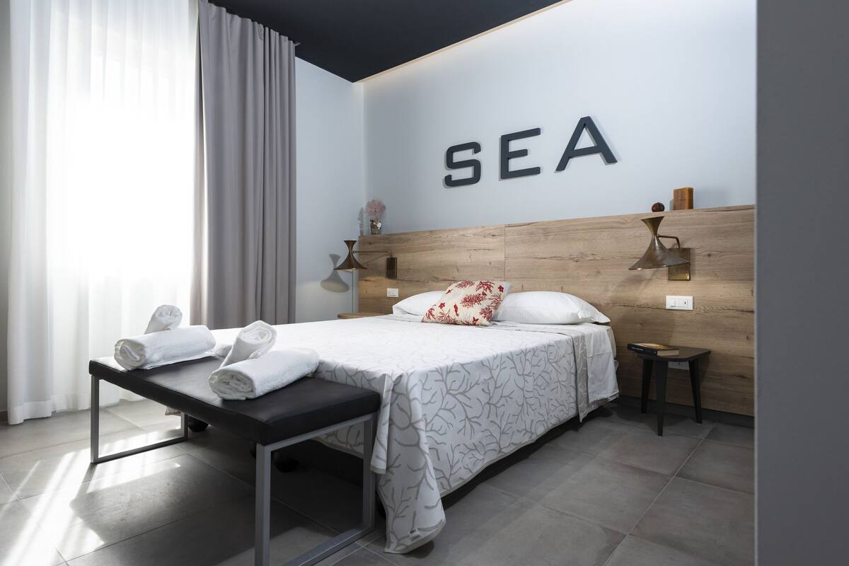 Quarto de hotel moderno com cama, mesa de cabeceira, um banco e prateleira na parede.
