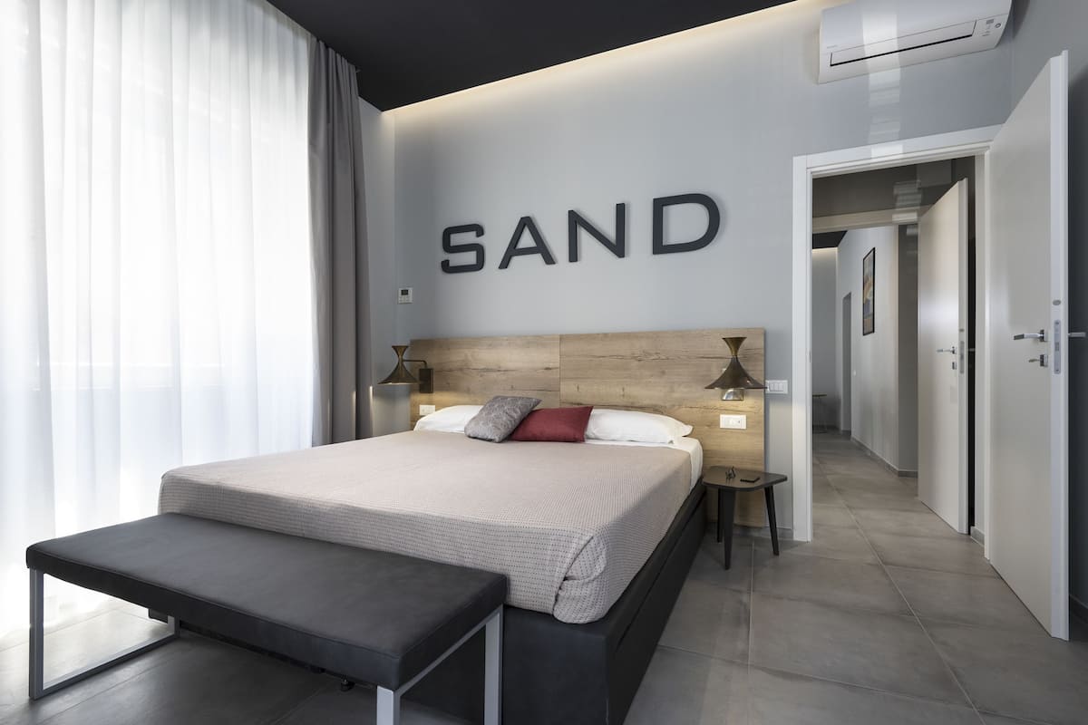 Quarto de hotel moderno com cama, mesas de cabeceira, um banco e um corredor que leva a uma porta.