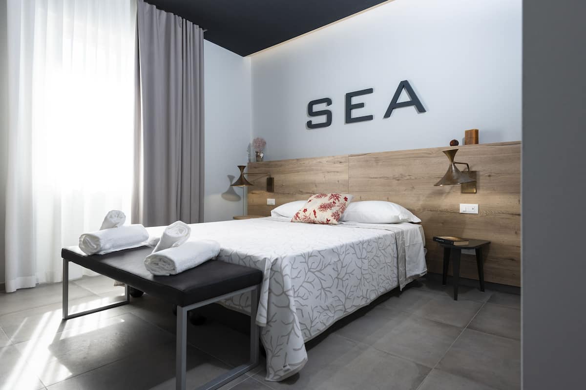 Quarto de hotel moderno com cama, um banquinho, mesa de cabeceira e parede com a palavra 'SEA'.