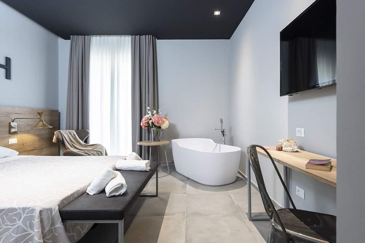 Quarto de hotel moderno com cama, área de estar, banheira e televisão presa na parede.