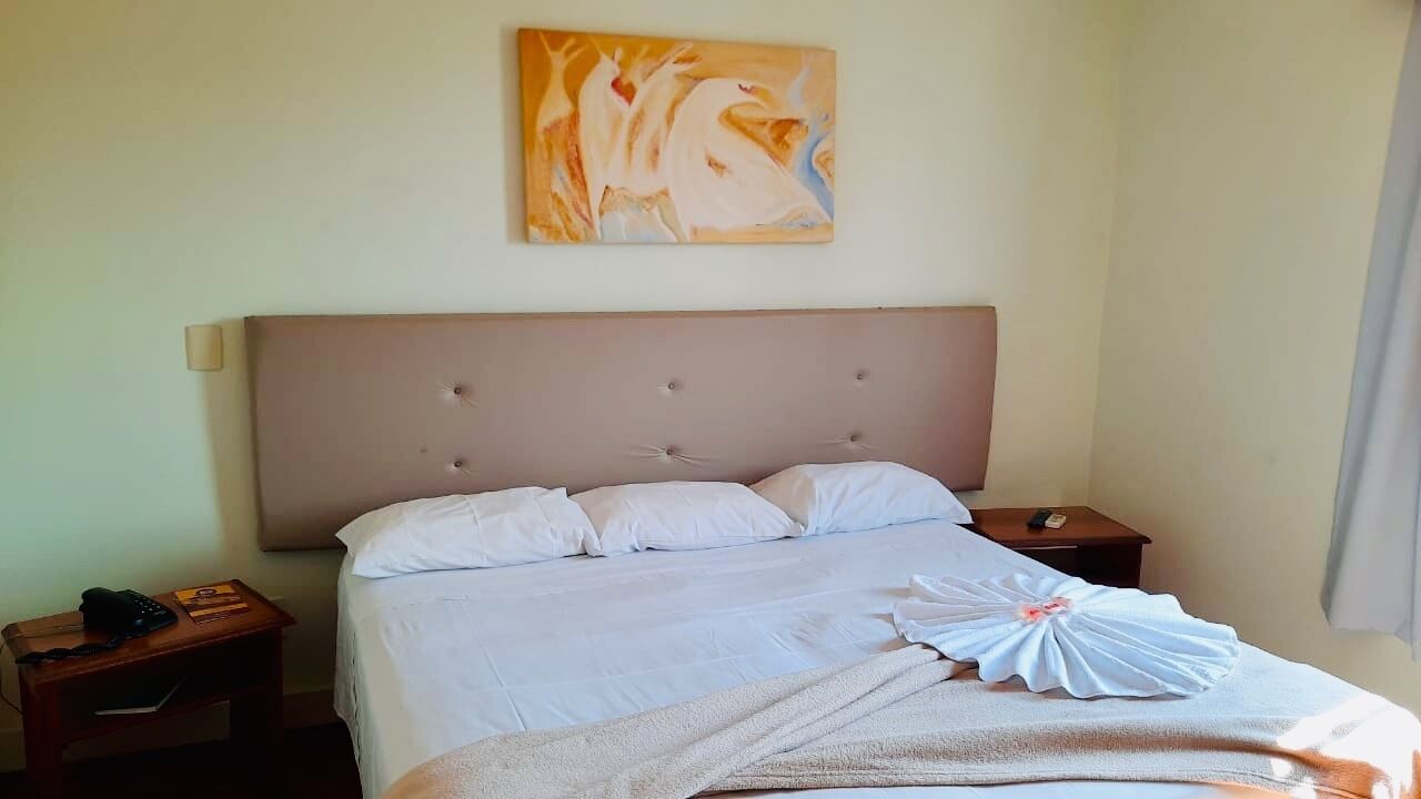 Cama bem arrumada com lençóis brancos e toalha decorativa, mesa de cabeceira com telefone e controle remoto, e um quadro na parede.