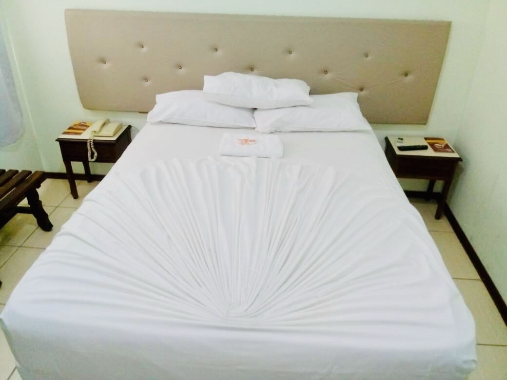 Uma cama bem arrumada com cabeceira estofada, duas mesas de cabeceira com um telefone e um livro, e um pequeno banco à esquerda.