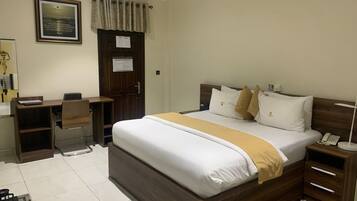 Quarto de hotel com cama, mesa de cabeceira, escrivaninha, cadeira e uma frase emoldurada na parede.