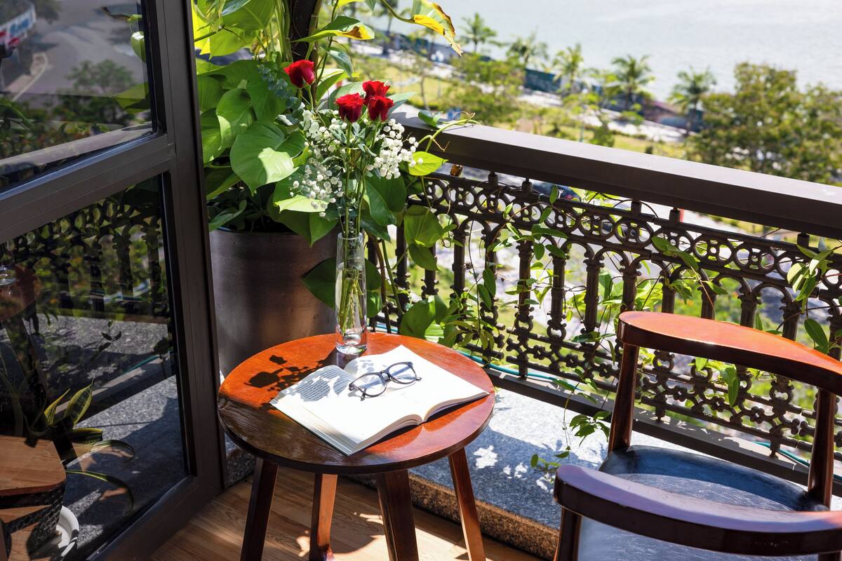 Uma varanda com uma mesa de madeira, um vaso com flores vermelhas e vista para o oceano e palmeiras.