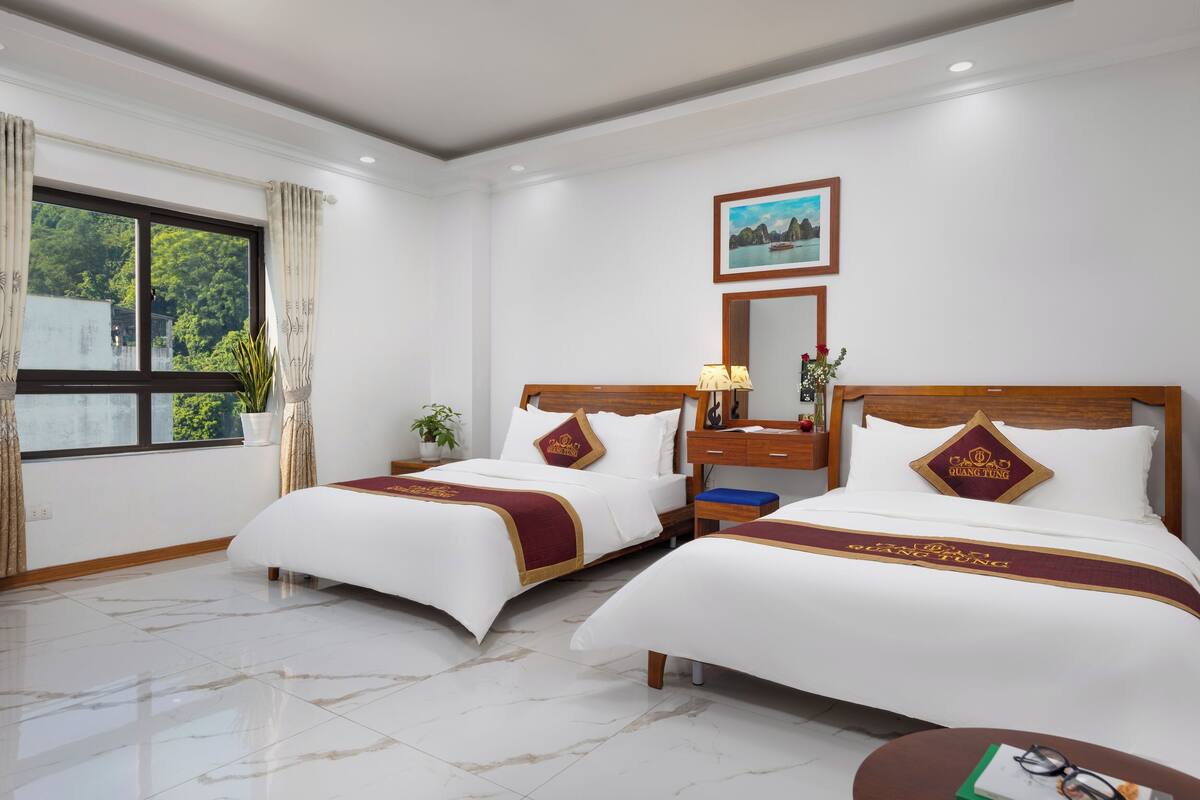 Quarto de hotel com duas camas, uma janela grande com cortinas, uma mesa de cabeceira com abajur e um quadro na parede.