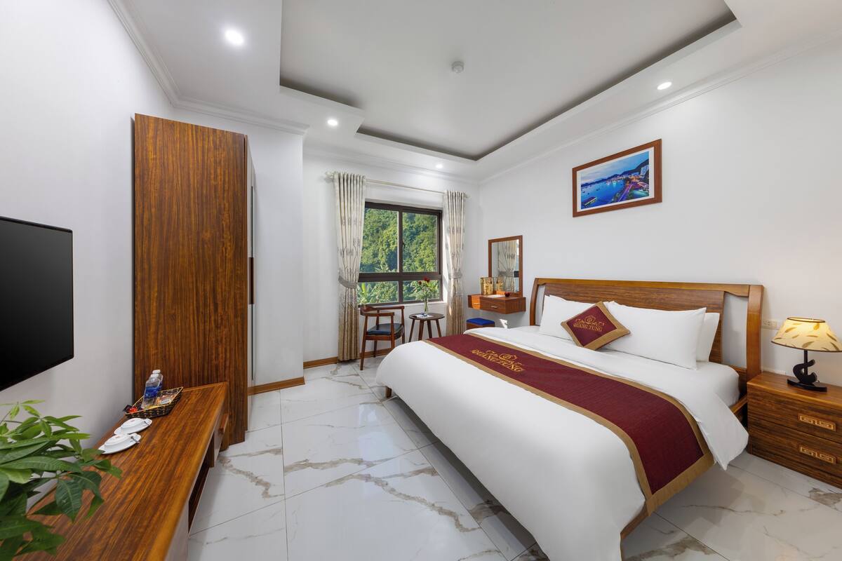 Quarto de hotel com uma cama grande, uma escrivaninha com cadeira, uma televisão e uma janela com cortinas.
