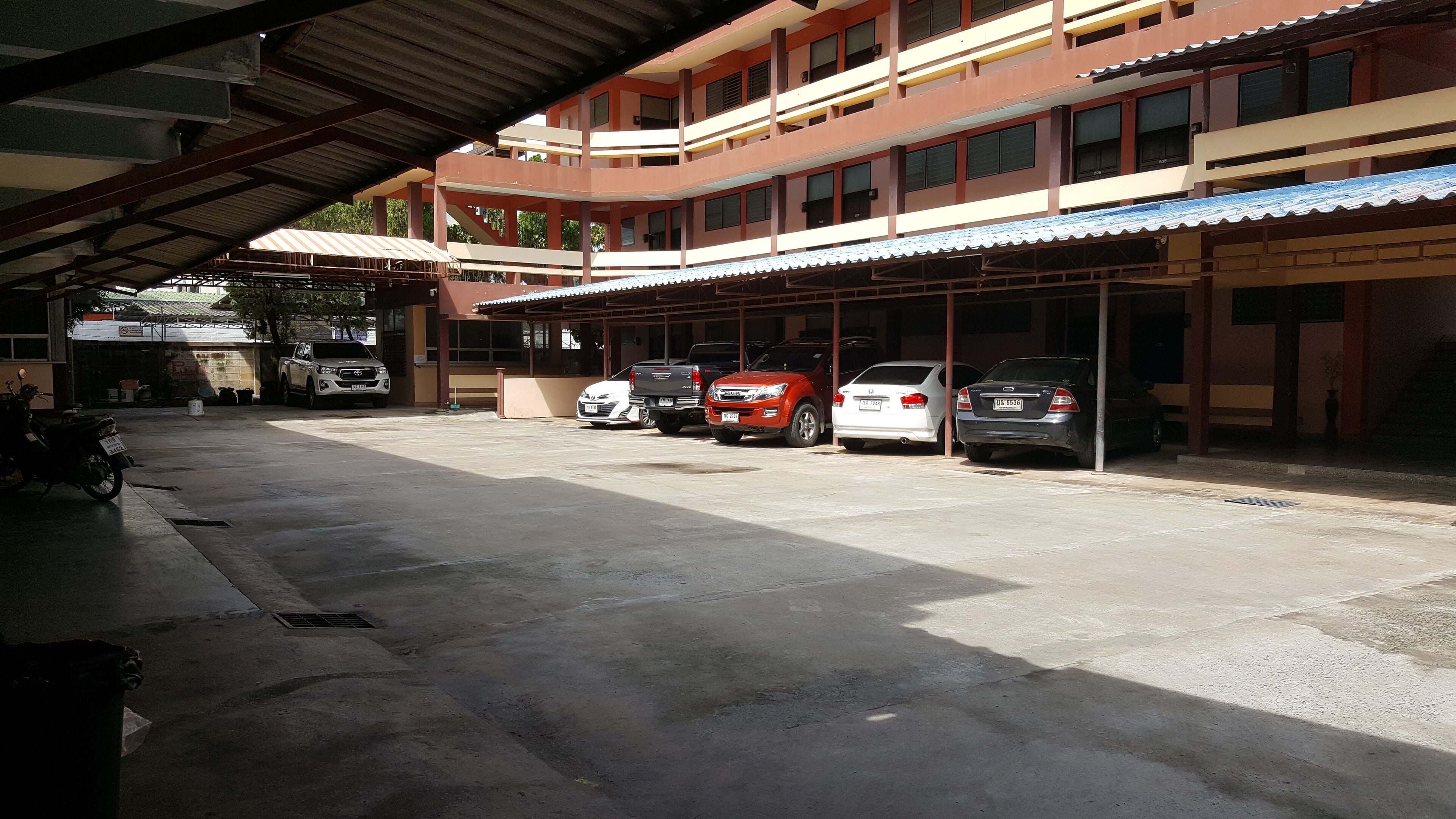 Uma área de estacionamento com vários carros e uma motocicleta, uma estrutura coberta e um edifício com vários andares e janelas.