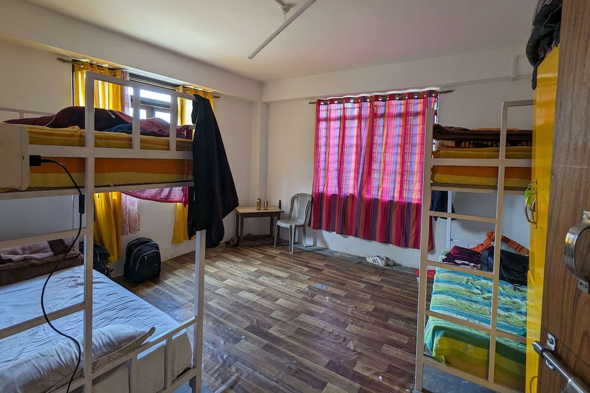 Quarto de dormitório com beliches, uma pequena mesa, uma cadeira e uma janela com cortinas.