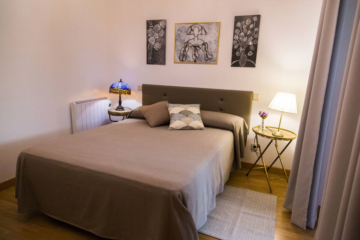 Quarto com cama grande, mesa de cabeceira com abajur e três quadros na parede.